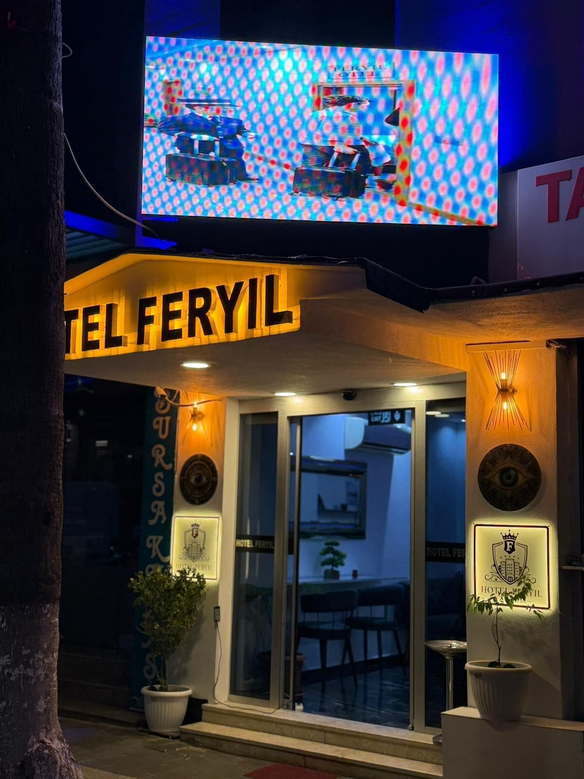 Entrada de hotel com letreiro de neon, porta de vidro e planta em vaso.