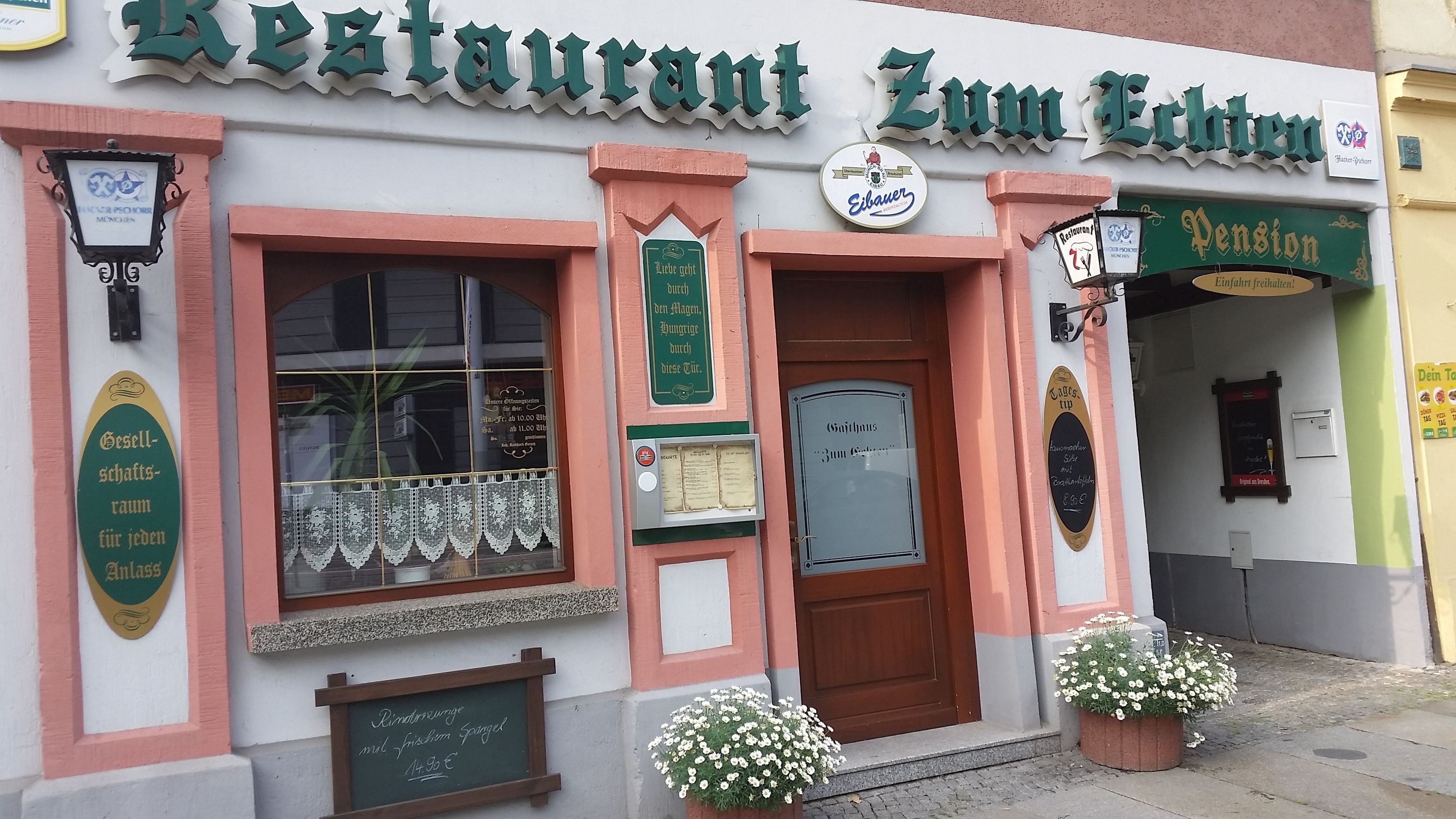 Um restaurante com fachada verde e branca, um letreiro com a inscrição "Zum Eichen" e um pequeno mostrador com flores.