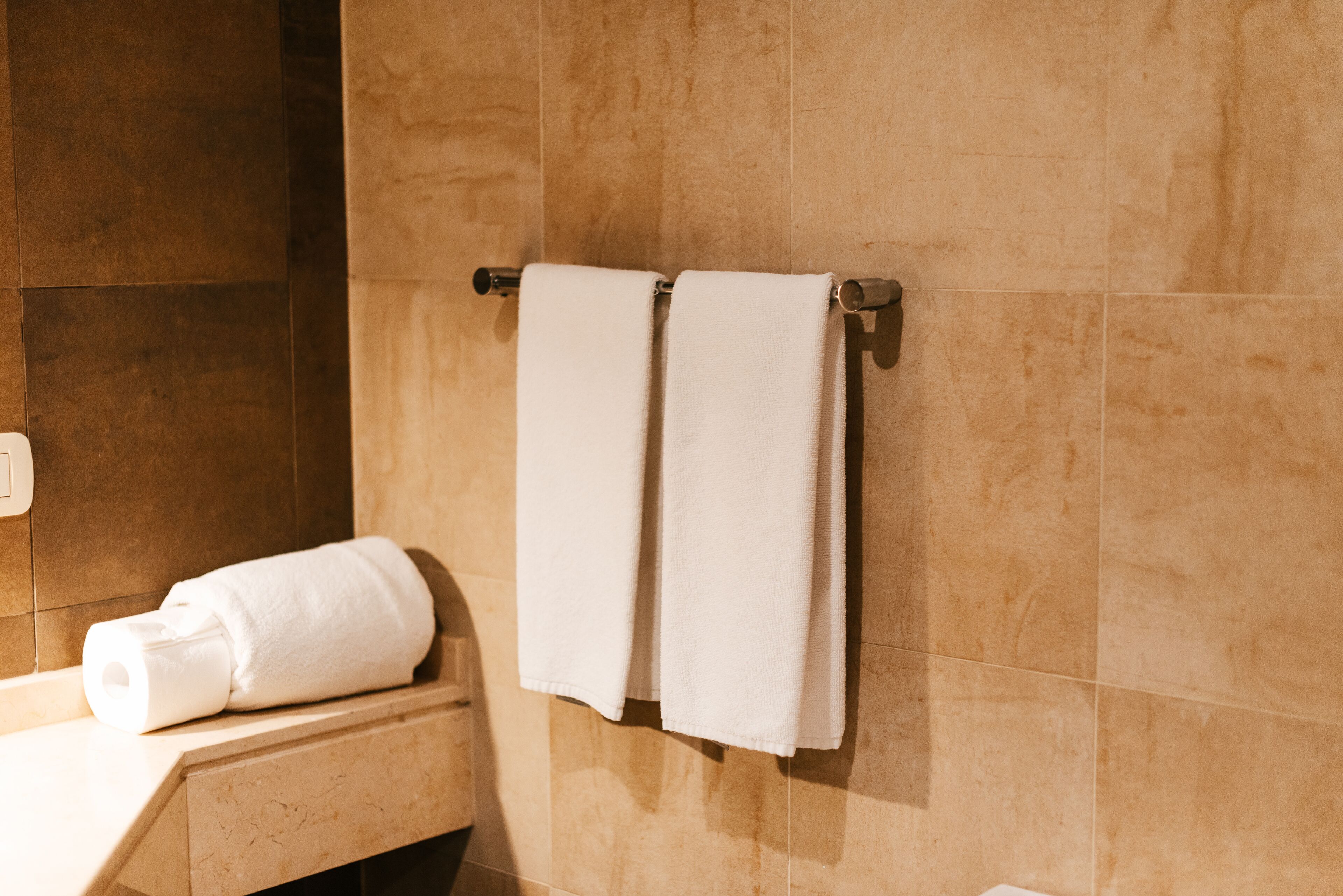 Casa de banho com azulejos bege, um porta-toalhas com duas toalhas e um rolo de papel higiénico no balcão.