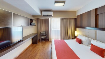 Quarto de hotel com uma cama, uma escrivaninha, uma cadeira, uma televisão e um ar condicionado.