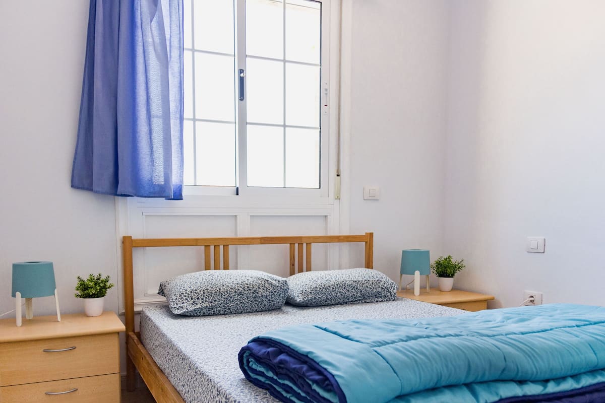 Quarto com cama de madeira, duas mesas de cabeceira, uma janela com cortina azul e uma pequena planta em cada mesa.