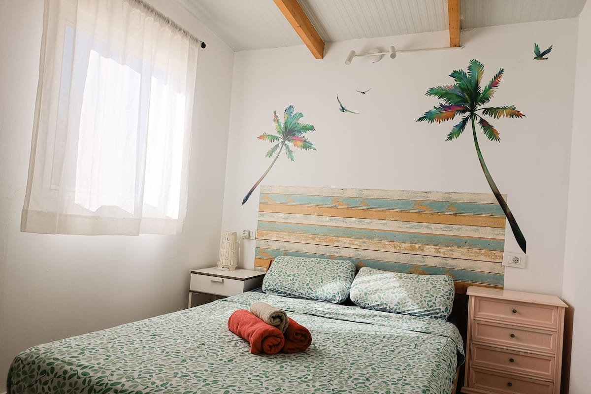 Quarto com cama, mesa de cabeceira, penteadeira, janela com cortinas leves e arte na parede com palmeiras e pássaros.