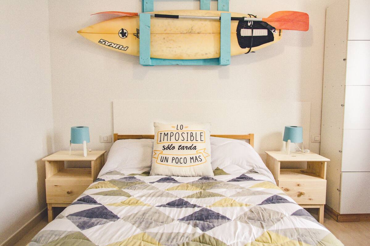 Quarto com cama, duas mesas de cabeceira, suporte para prancha de surf na parede e almofada com texto.