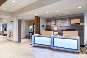 Uma área de recepção de hotel com design moderno, destacando um balcão de recepção com painéis iluminados, uma placa fixada na parede da Holiday Inn Express e uma geladeira ao fundo.