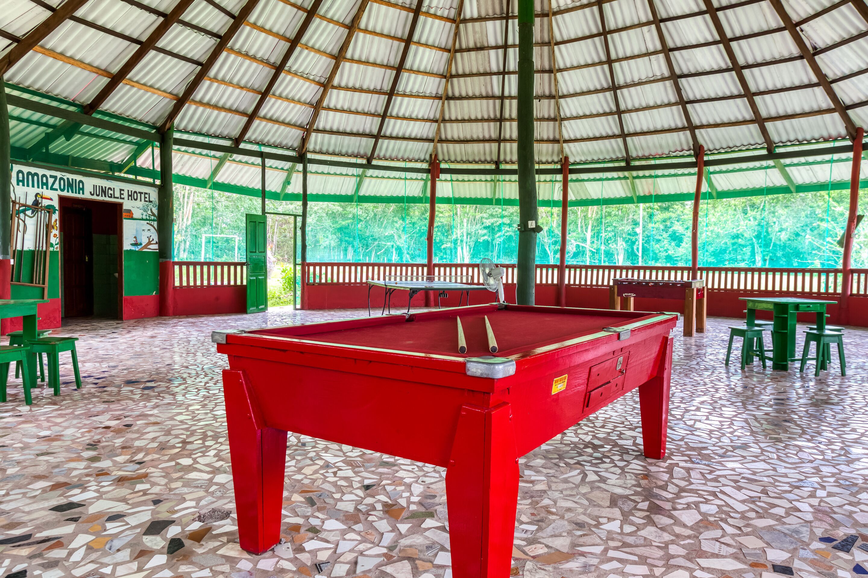 Mesa de bilhar numa área coberta, com esquema de cores verde e vermelho, rodeada por cadeiras verdes e uma mesa.