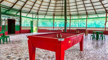 Mesa de bilhar numa área coberta, com esquema de cores verde e vermelho, rodeada por cadeiras verdes e uma mesa.