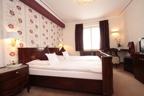 Quarto de hotel com uma cama grande, duas mesas de cabeceira, uma escrivaninha, uma cadeira, uma televisão e uma janela com cortinas.