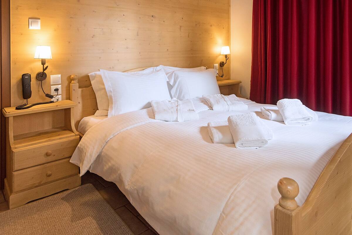 Quarto de hotel com cabeceira de madeira, duas mesas de cabeceira com luminárias, uma cortina vermelha e toalhas dobradas na cama.