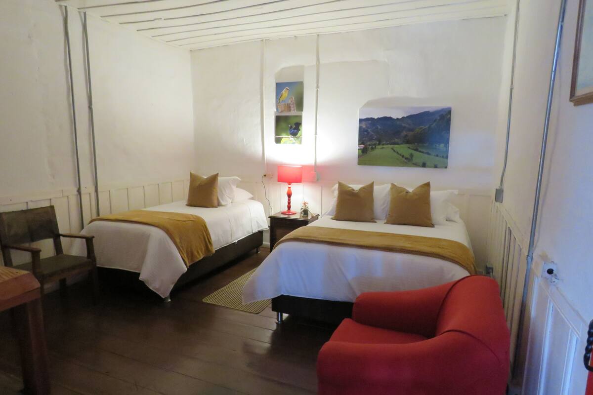 Quarto de hotel com duas camas, uma cadeira vermelha, uma mesa de cabeceira com abajur e um quadro na parede.