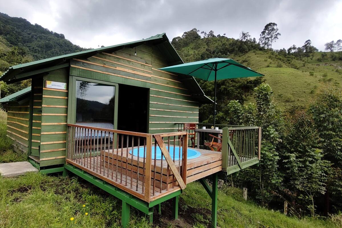 Uma cabana pequena e verde com um alpendre de madeira e piscina, rodeada por vegetação exuberante e colinas.