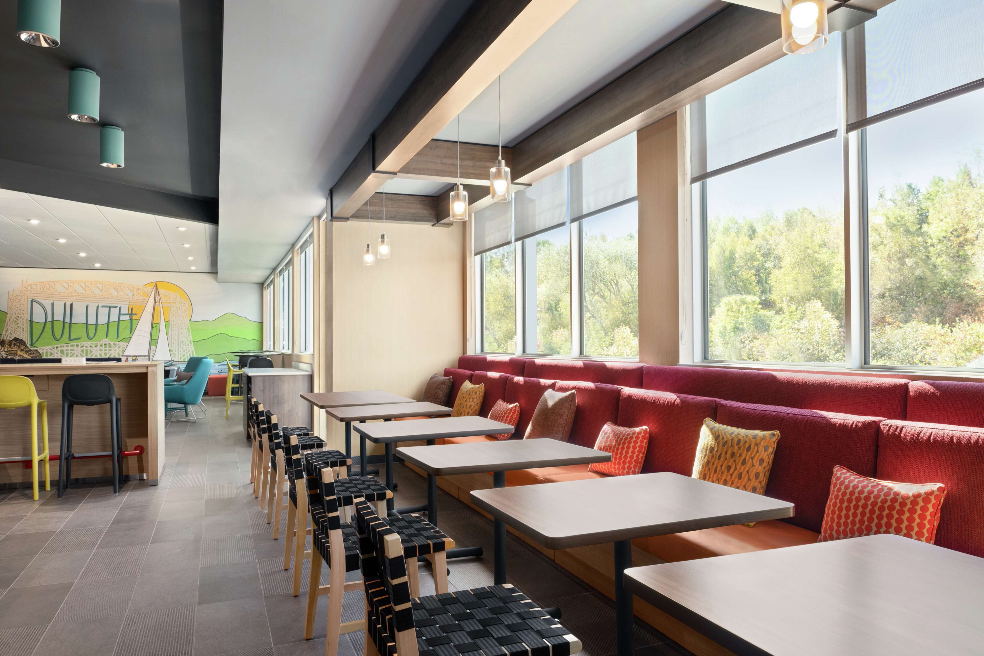 Interior de restaurante moderno com mesas e bancos altos, grandes janelas com vista para o verde e um mural na parede.