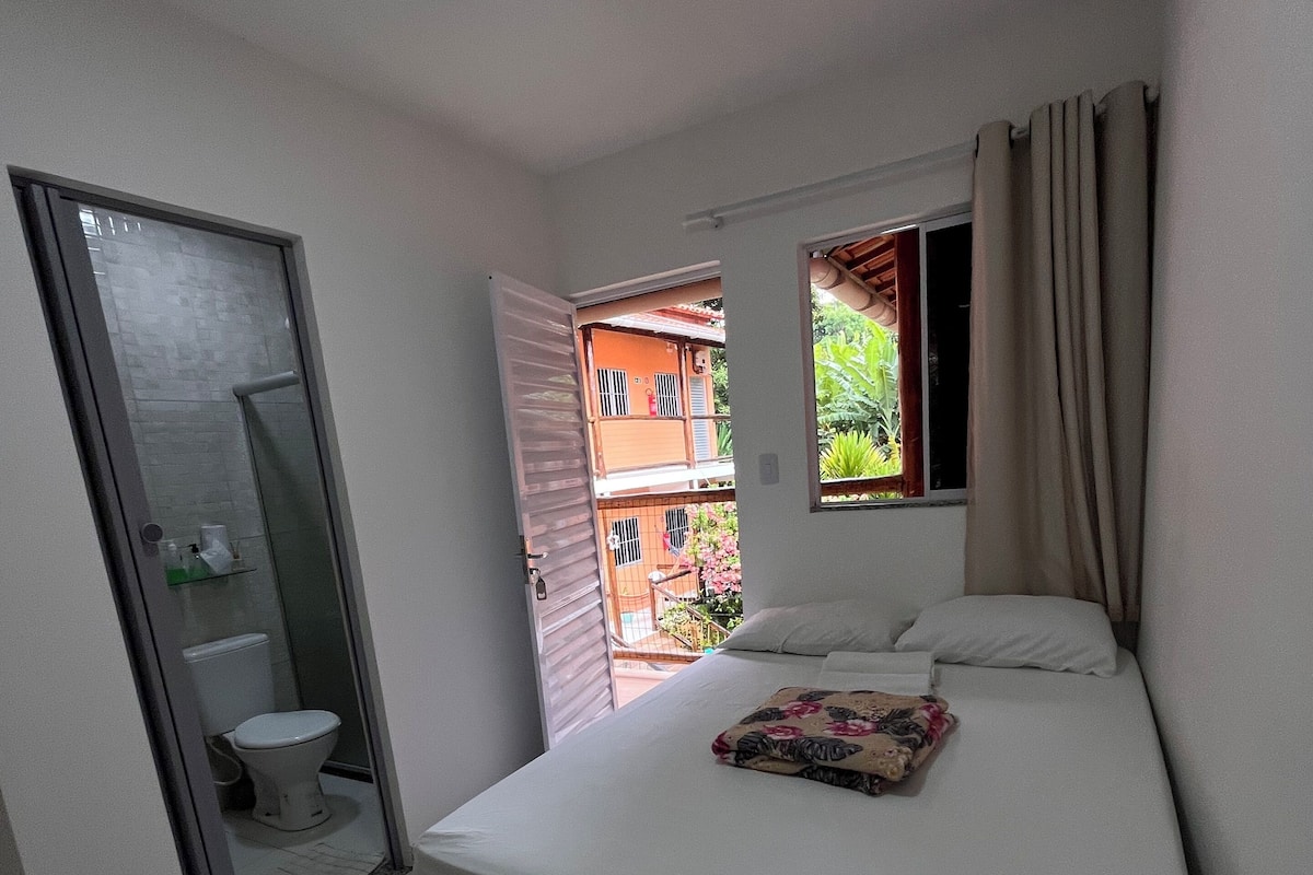 Um quarto com cama, banheiro com vaso sanitário e chuveiro, e varanda com plantas.