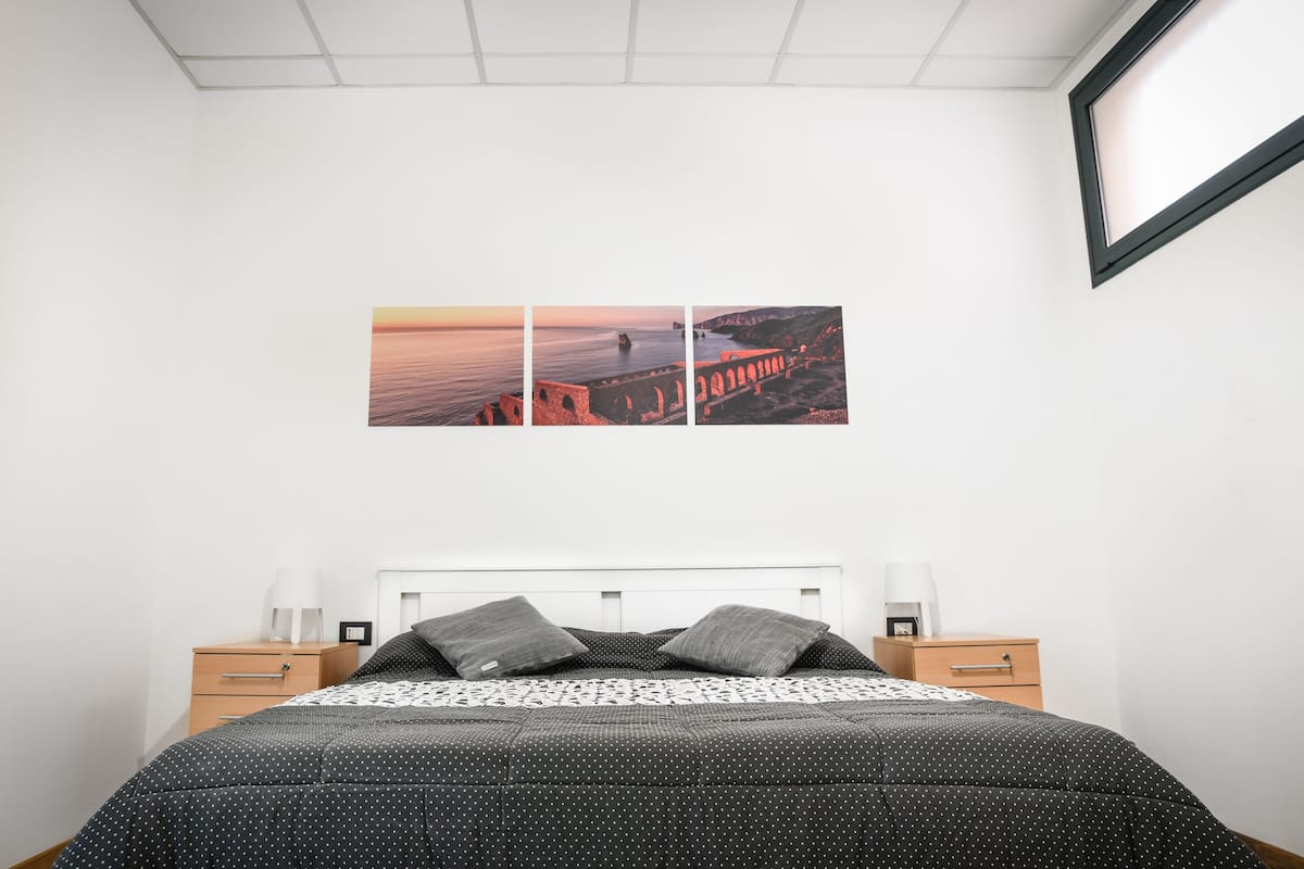 Quarto com cama, duas mesas de cabeceira, um criado-mudo e um quadro na parede retratando um pôr do sol sobre uma ponte.
