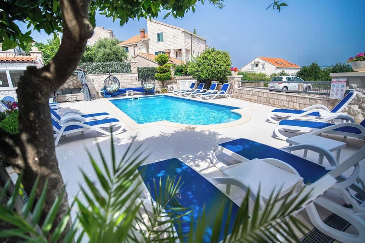 Piscina com espreguiçadeiras, parede de pedra e vista para casas ao fundo.