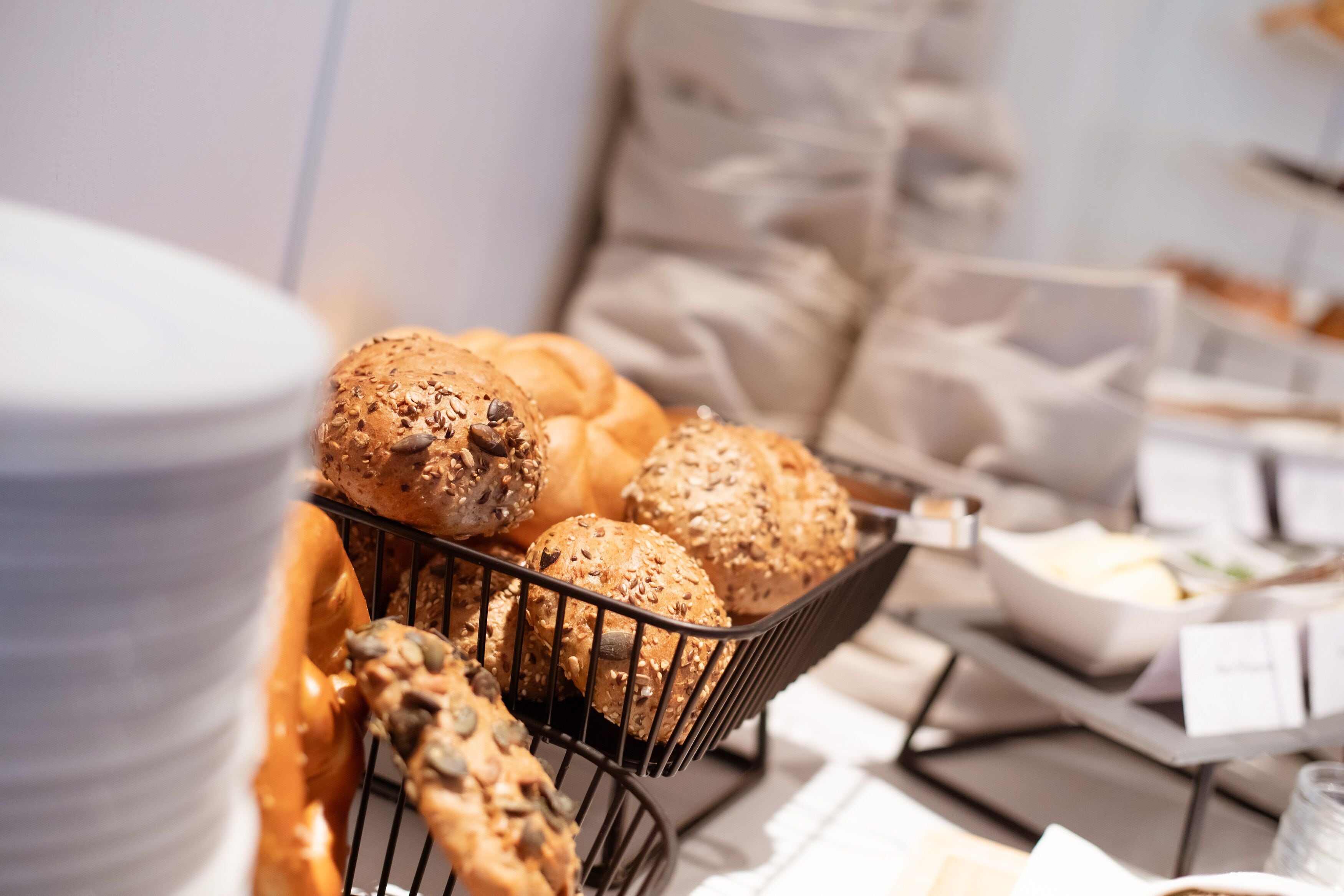 Uma cesta com diversos produtos de panificação, incluindo pães e muffins, sobre uma mesa com toalha branca.