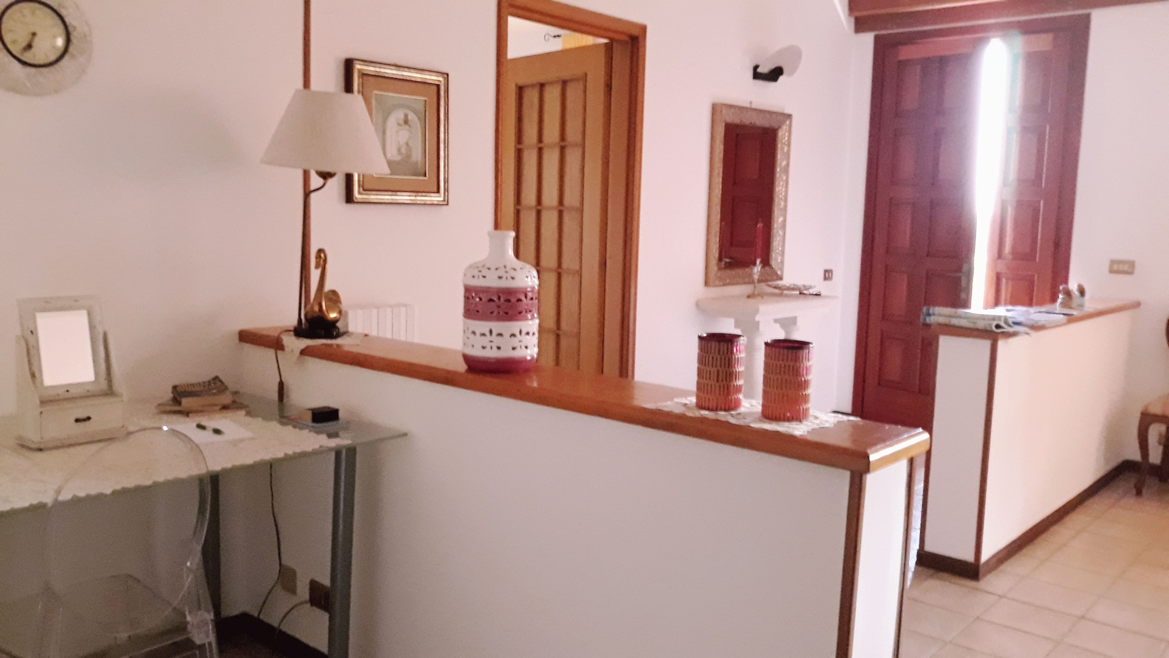 Uma cozinha compacta com bancada de madeira, pia branca e espelho acima. Há uma moldura na parede e uma porta ao fundo.