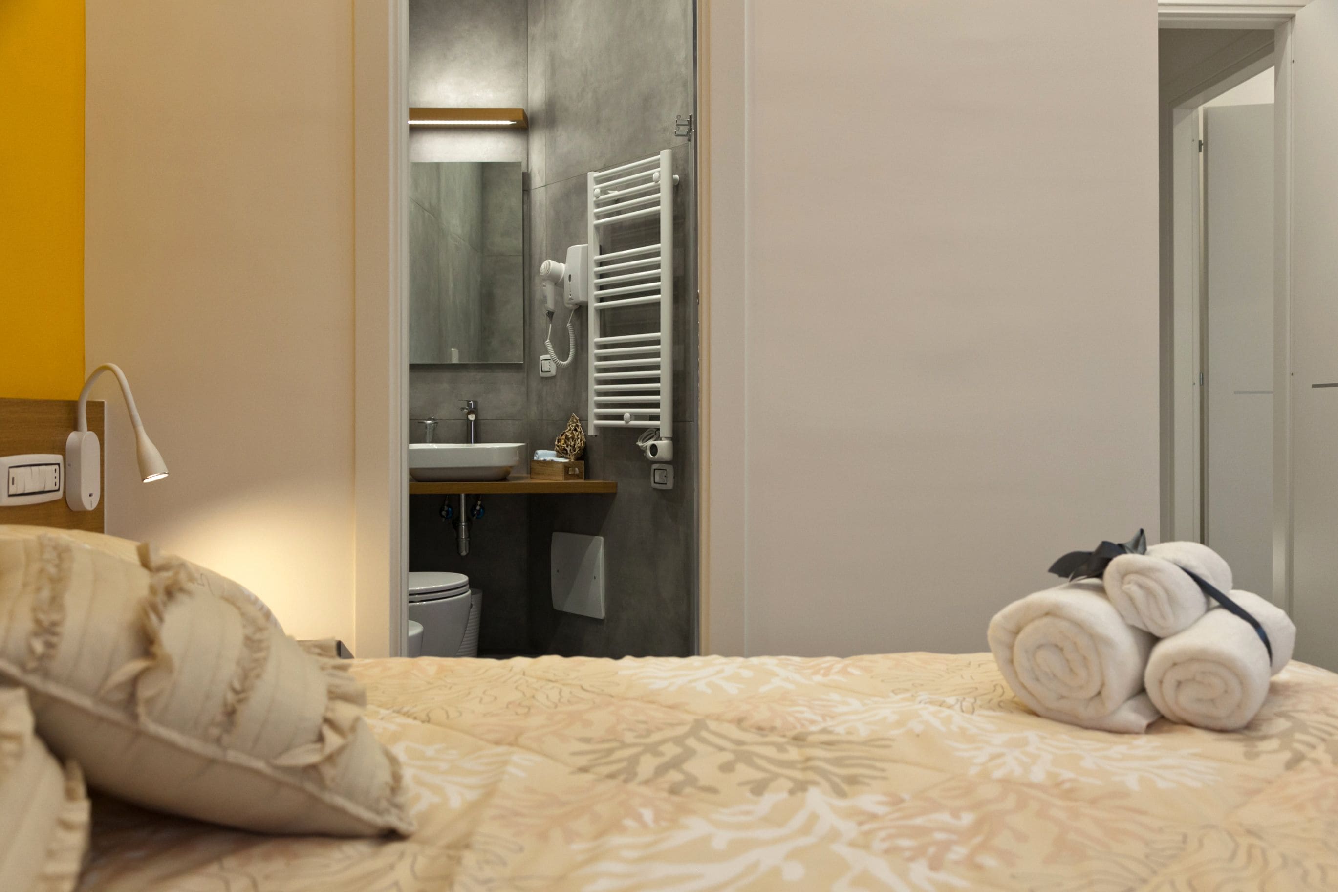Quarto de hotel com cama, luminária, porta-toalhas e banheiro visível por uma porta aberta.