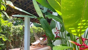Um caminho de jardim leva a um edifício branco com telhado de madeira, cercado por vegetação exuberante e uma rede.