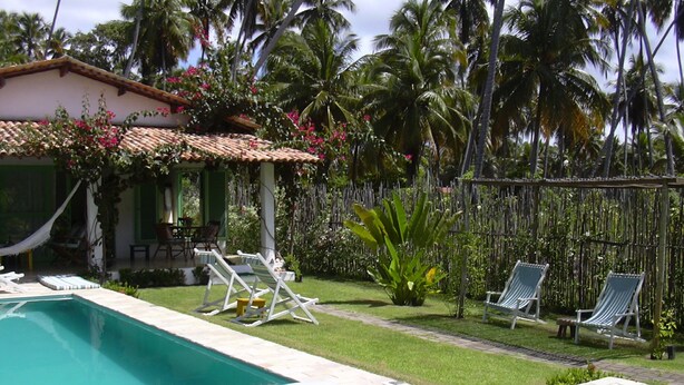 Piscina com espreguiçadeiras e boia inflável, cercada por um jardim com palmeiras e uma casa com telhado ladrilhado.