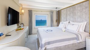Quarto de hotel com uma cama, escrivaninha, televisão, janela com vista para o mar e uma luminária de teto.