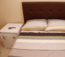 Uma cama com cabeceira marrom, duas almofadas e um edredom listrado. Uma mesa de cabeceira branca com um controle remoto e um filtro de tomada é visível.