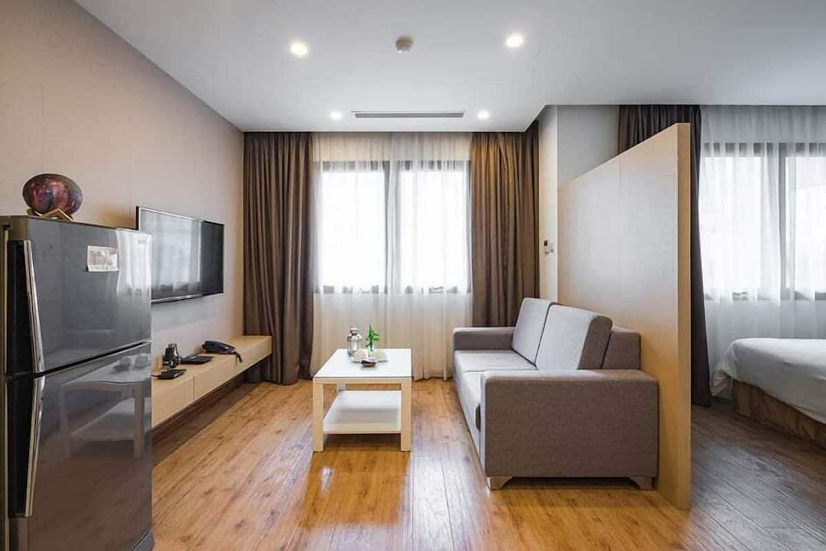 Quarto de hotel moderno com piso de madeira, sofá, cama, televisão, frigorífico e uma pequena mesa com objeto decorativo.