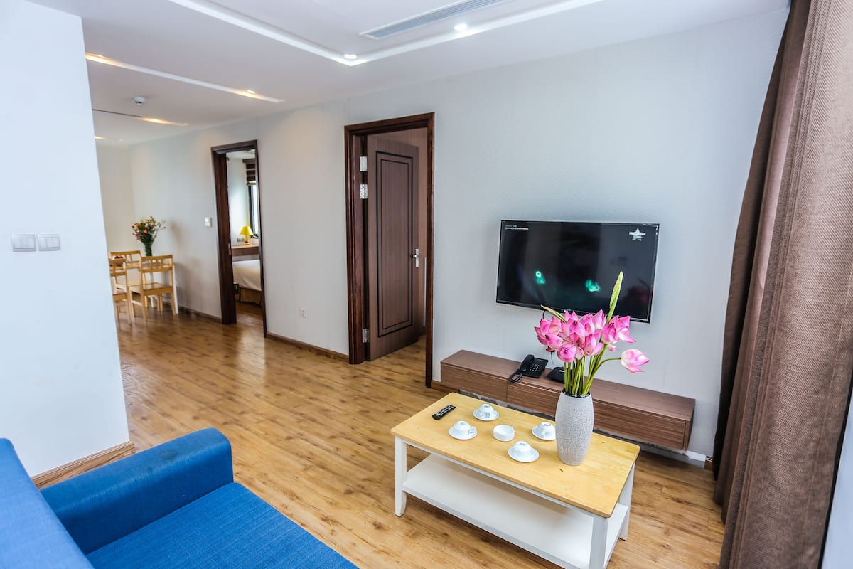 Uma sala de estar moderna com piso de madeira, um sofá azul, uma mesa de centro com um vaso de flores, uma TV de tela plana e um banco de madeira.