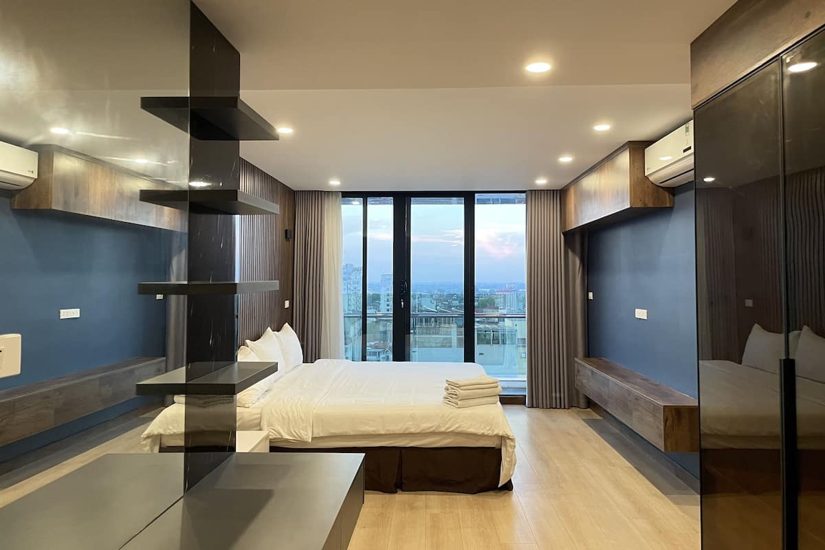 Quarto de hotel moderno com uma cama grande, uma escrivaninha e vista para a cidade através de janelas do chão ao teto.