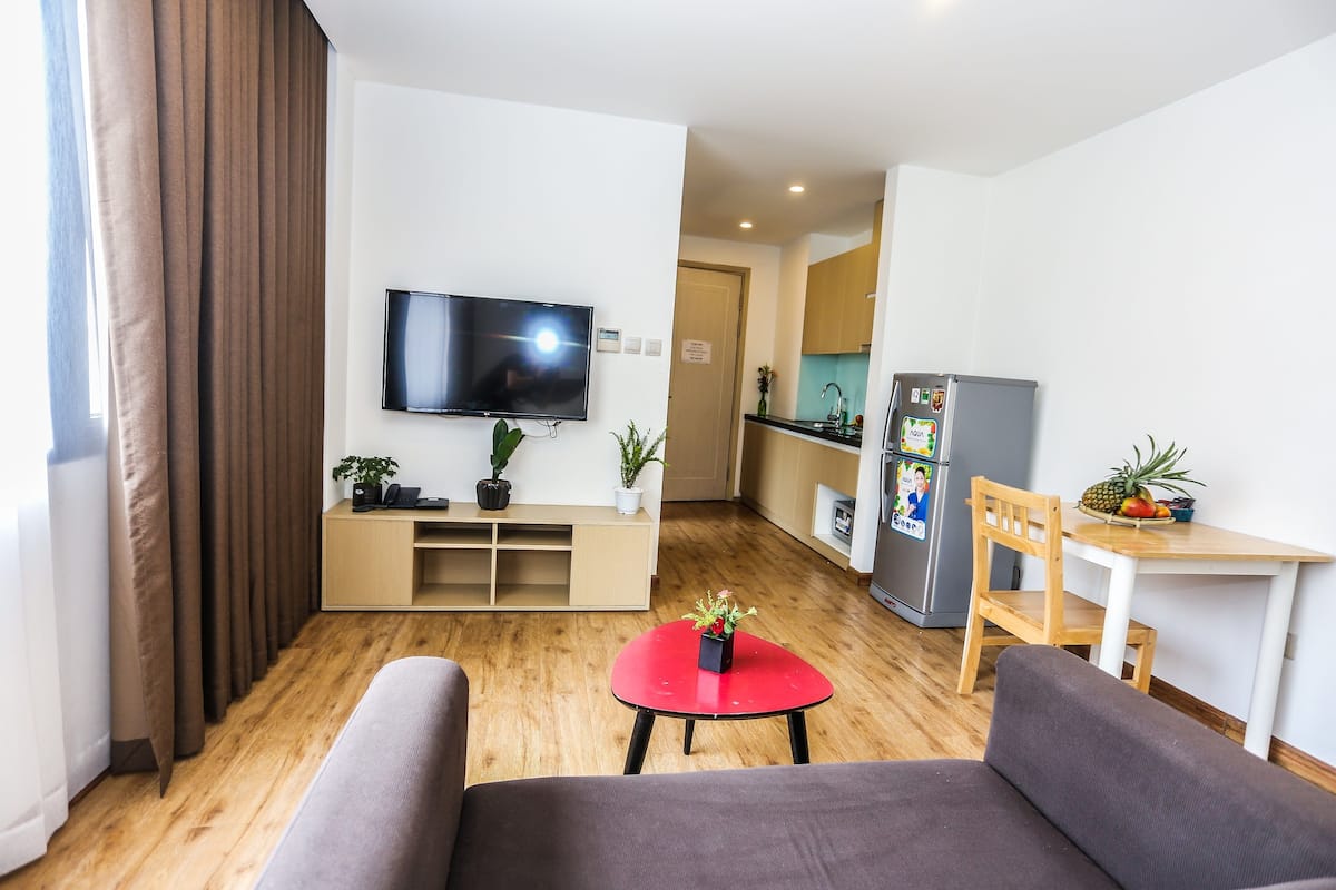 Uma sala de estar moderna com uma televisão de tela plana, uma mesa vermelha e um sofá. Uma cozinha é visível ao fundo, com uma geladeira e um espaço de refeições.