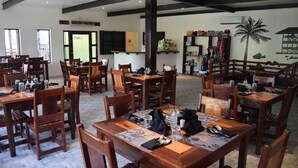 Um restaurante com mesas e cadeiras de madeira, um pilar grande e uma parede com um mural de palmeira.