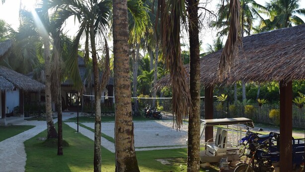 Um resort tropical com palmeiras, uma construção de palha e um jipe estacionado em primeiro plano.
