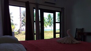 Quarto de hotel com uma cama, vista para palmeiras e praia, e uma cadeira perto da janela.