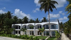 Um edifício moderno de dois andares com fachada branca, várias varandas e cercado por vegetação exuberante e palmeiras.