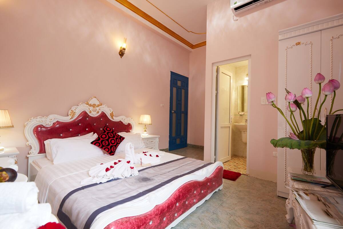 Um quarto com uma cama grande, uma porta azul, um espelho e um vaso com flores.