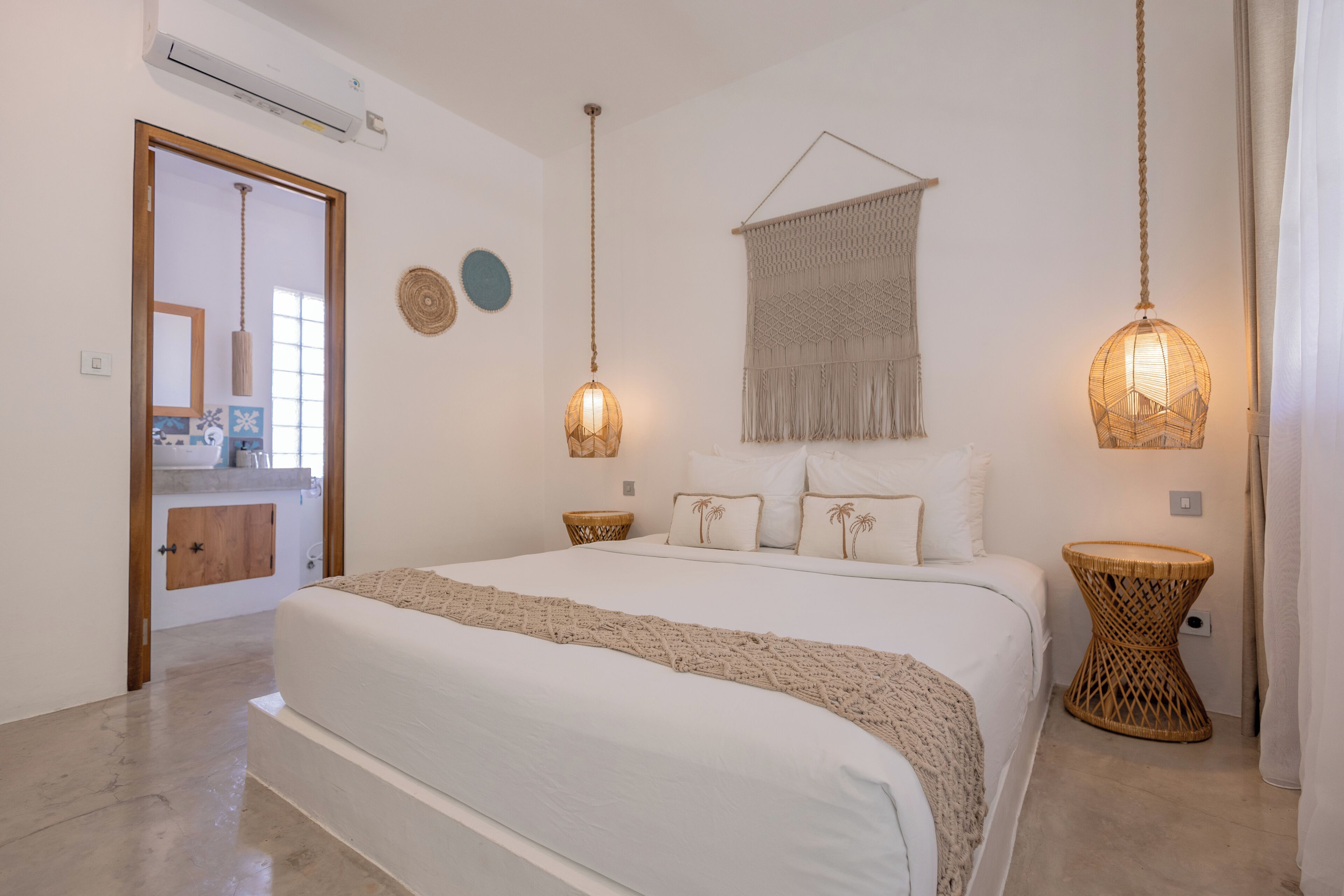 Quarto com uma cama grande, duas mesas de cabeceira, um lustre pendente, um macramê na parede e uma porta para o banheiro.