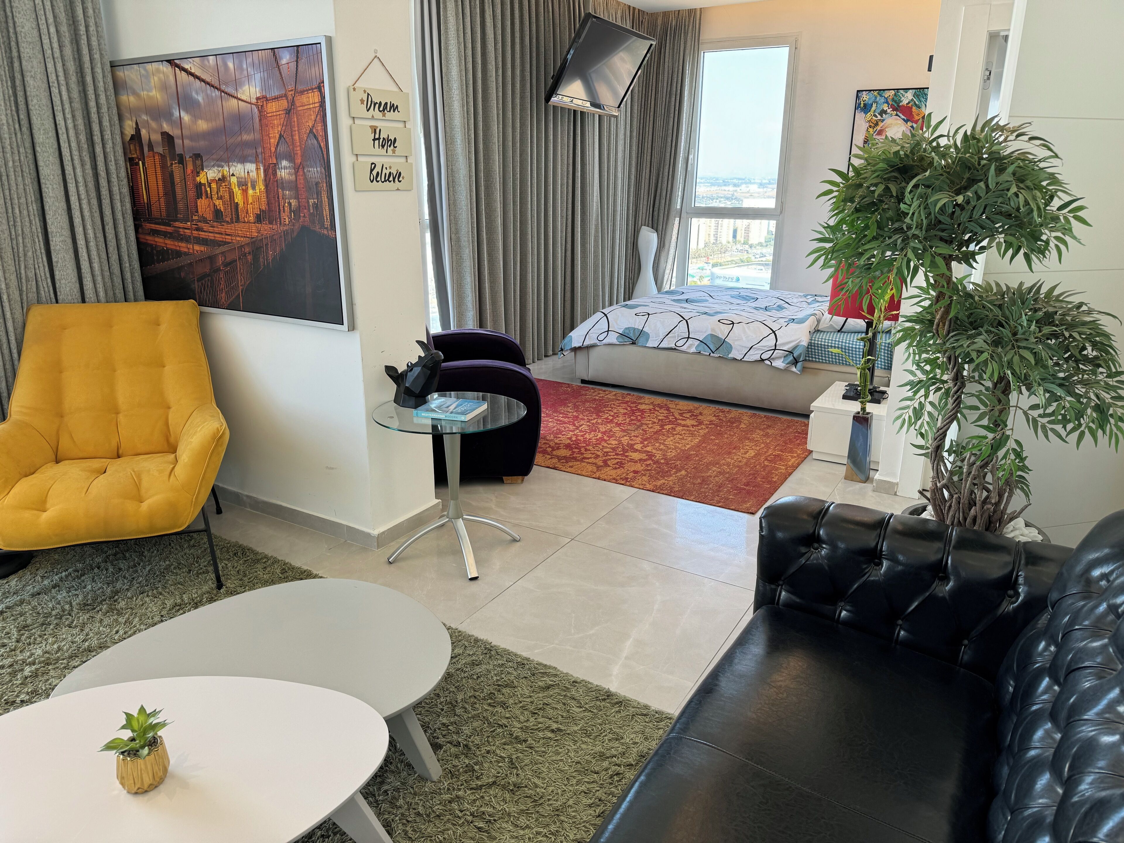 Uma sala de estar moderna com uma poltrona amarela, um sofá de couro preto, uma mesa de centro de vidro e uma pequena mesa branca com uma planta. Há uma cama com colcha estampada e uma janela grande com cortinas.