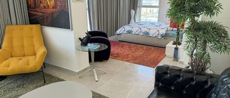 Uma sala de estar moderna com uma poltrona amarela, um sofá de couro preto, uma mesa de centro de vidro e uma pequena mesa branca com uma planta. Há uma cama com colcha estampada e uma janela grande com cortinas.