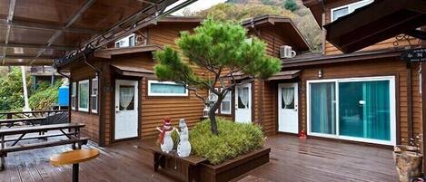 Uma cabana de madeira com varanda coberta, um pequeno jardim com uma árvore e um banco decorado com enfeites de boneco de neve.