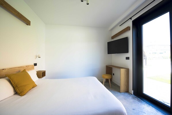Quarto moderno com uma cama grande, um banco de madeira, um banquinho pequeno e uma TV fixada na parede.