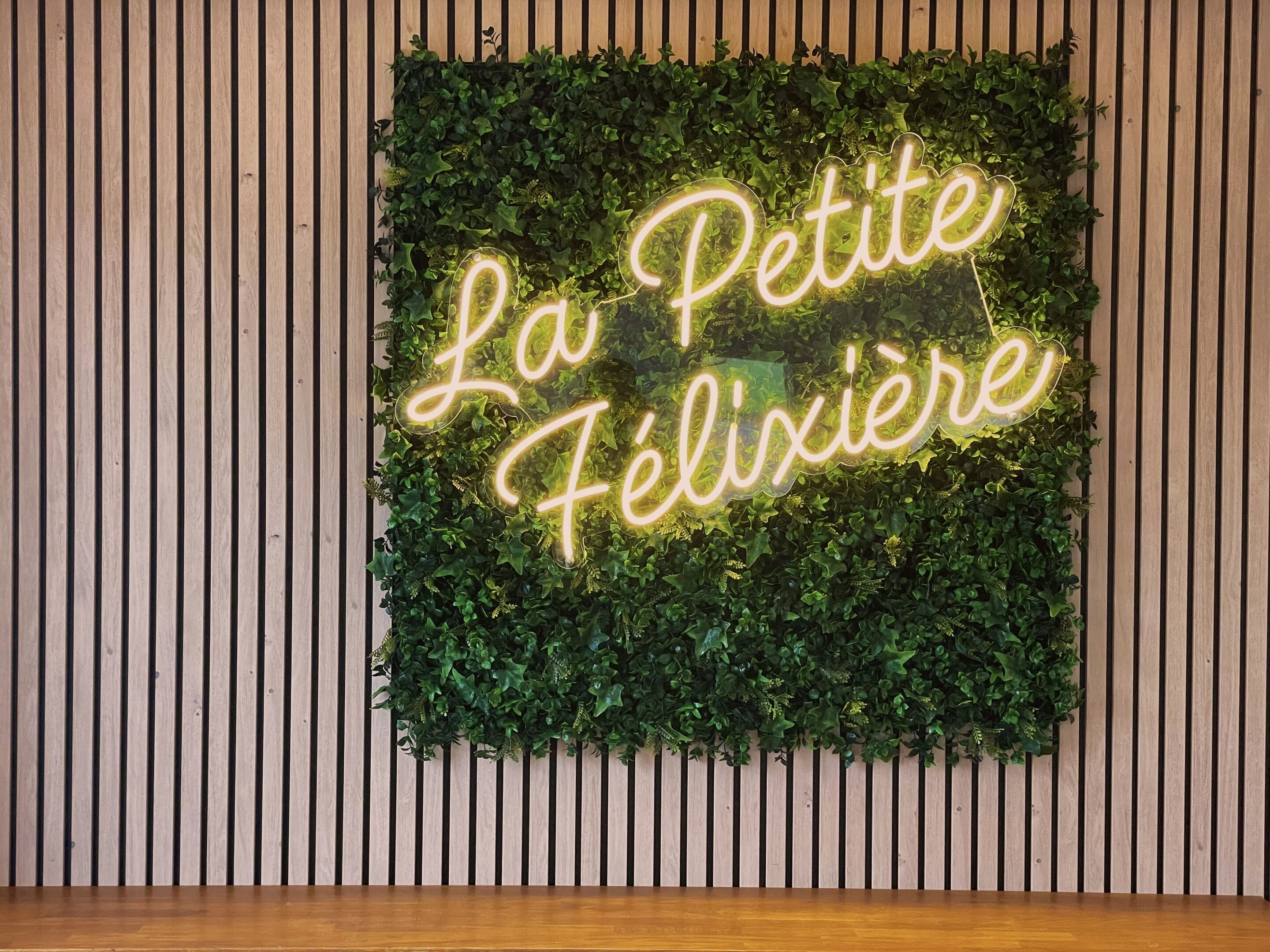 Um letreiro de neon com o texto 'La Petite Félixière' em frente a um painel de ripas de madeira vertical e uma parede de folhagem verde.