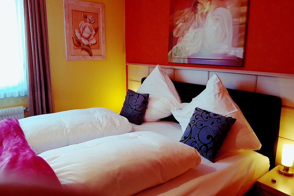 Quarto de hotel com uma cama, almofadas, mesa de cabeceira e abajur.