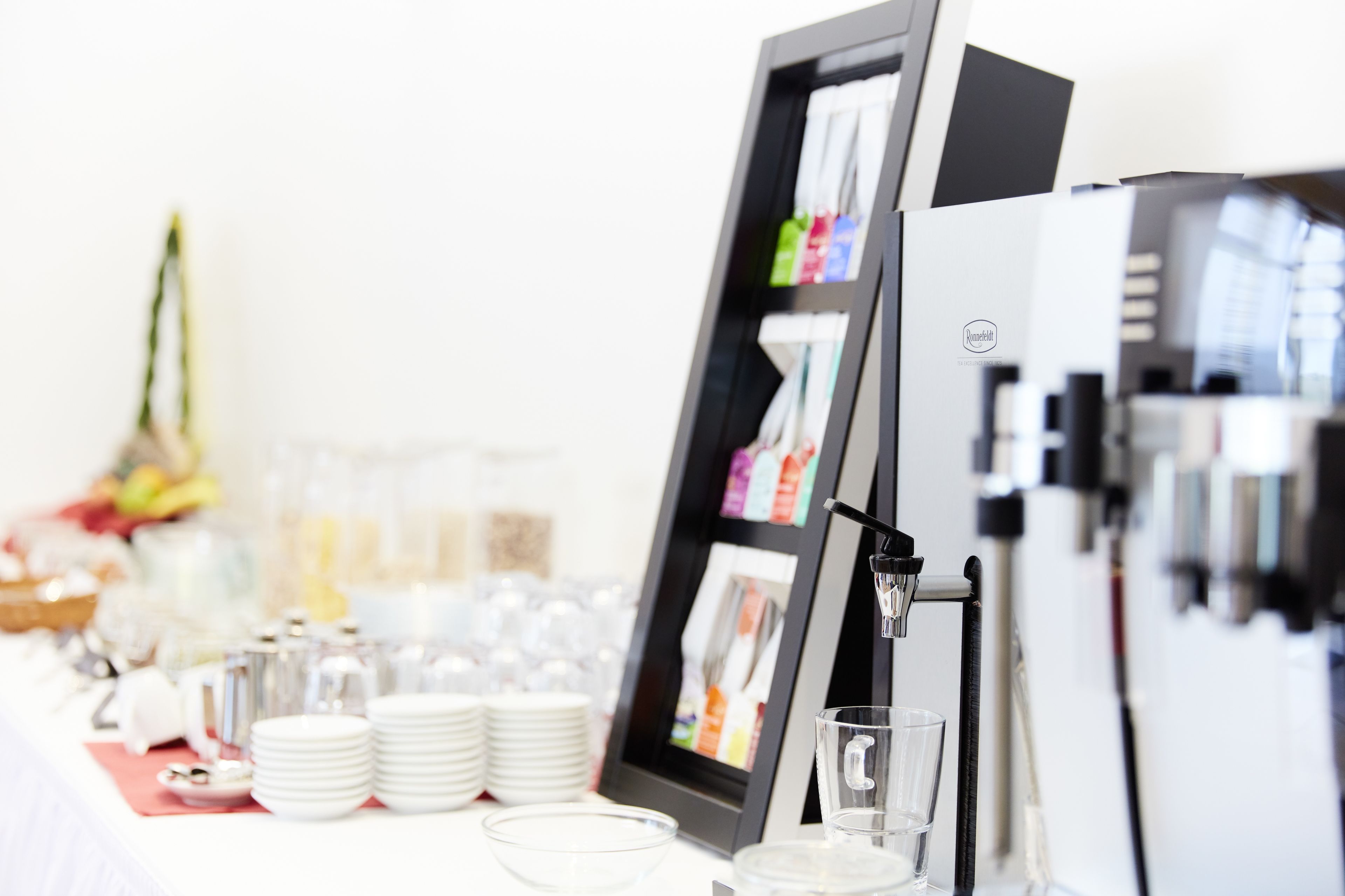 Uma mesa de buffet com dispensador de bebidas, pratos empilhados e um expositor de vidro.