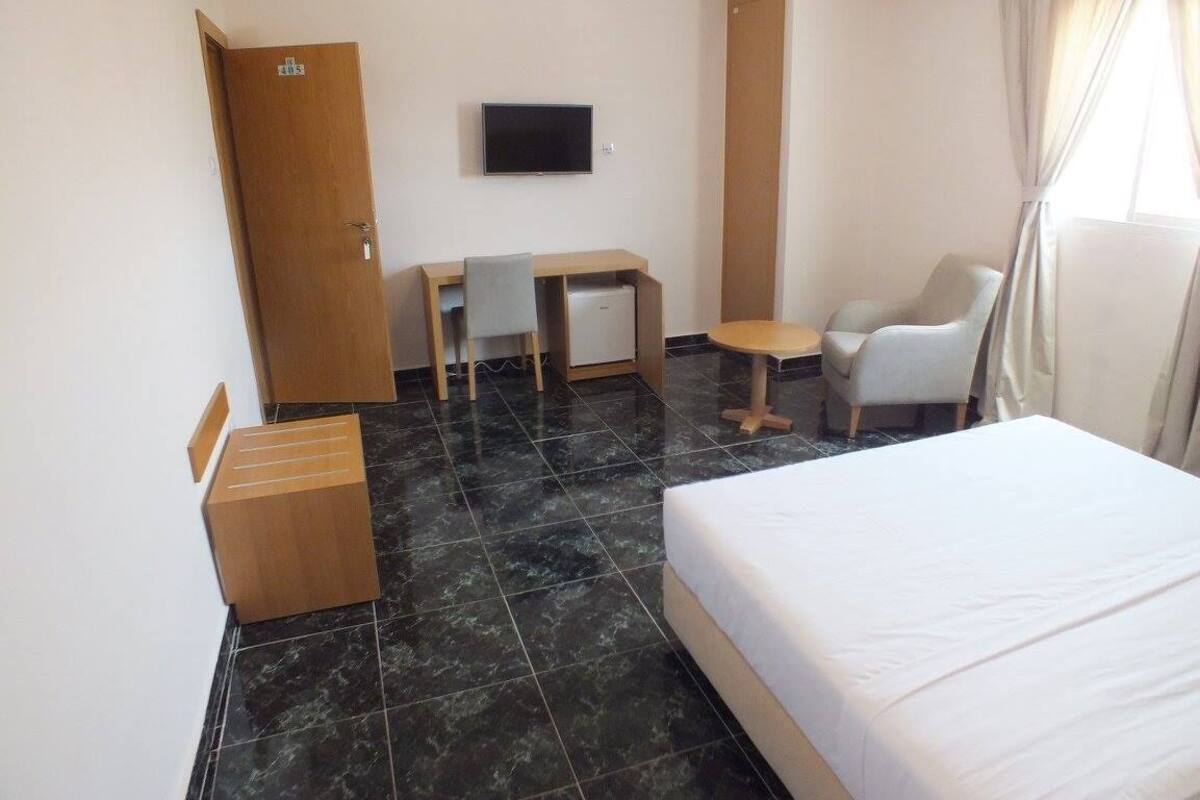 Quarto de hotel com cama, escrivaninha, cadeira, televisão e uma mesinha.