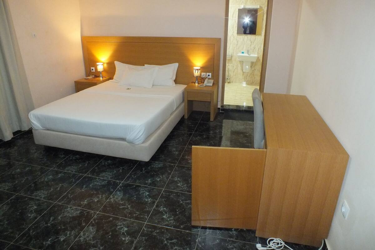 Quarto de hotel com cama, luminárias de cabeceira, mesa de cabeceira, espelho e escrivaninha de madeira.