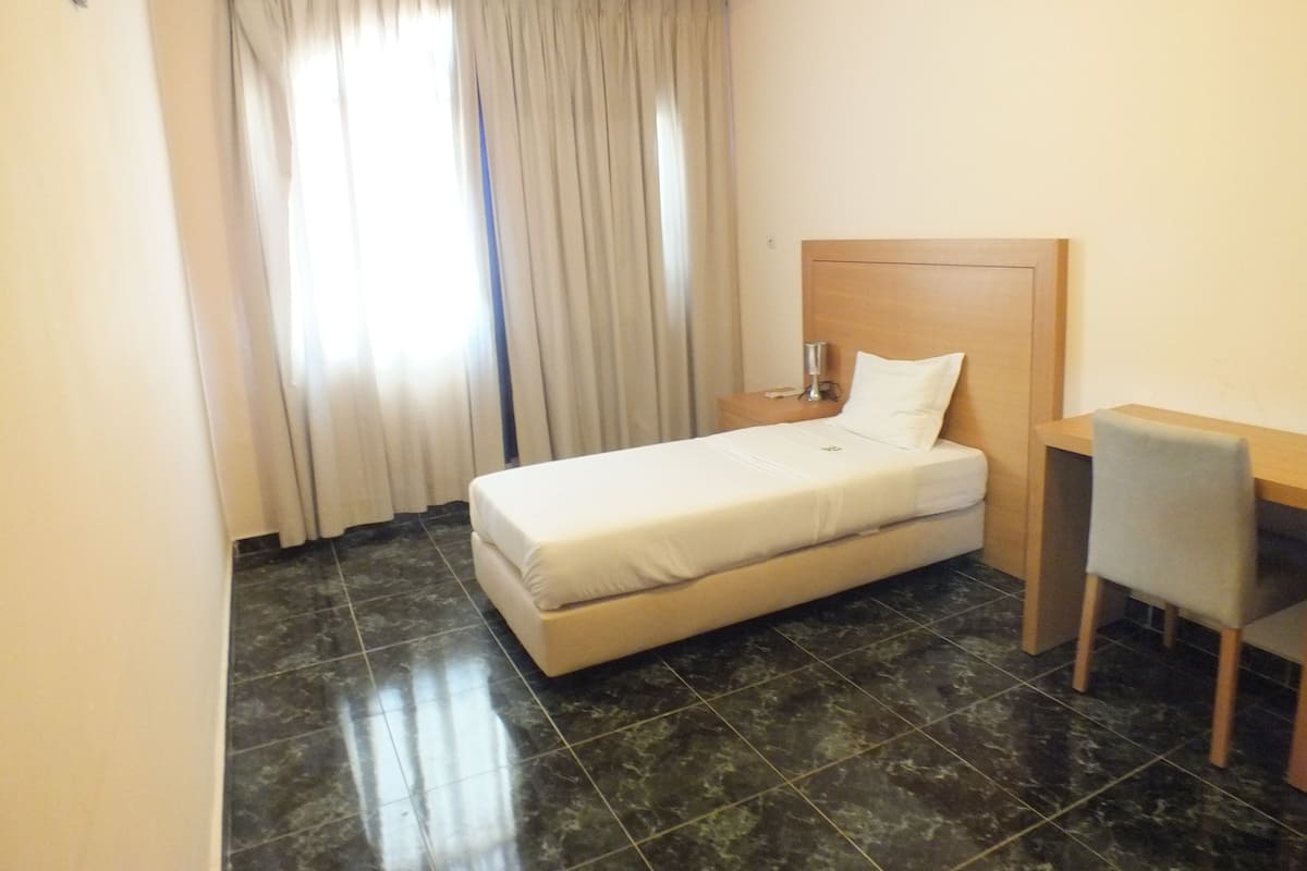 Quarto de hotel com cama, escrivaninha, cadeira, mesa de cabeceira e janela com cortinas.