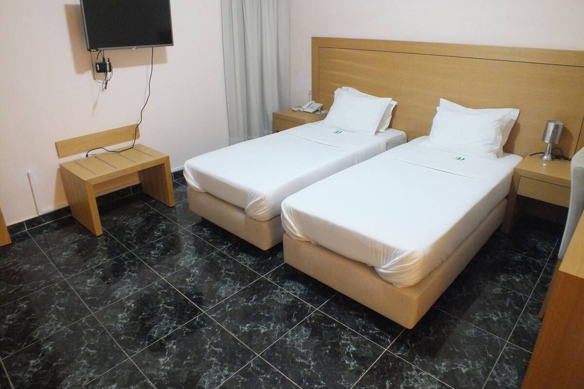 Quarto de hotel com duas camas de solteiro, televisão fixada na parede, mesa de cabeceira com abajur e um banco de madeira.