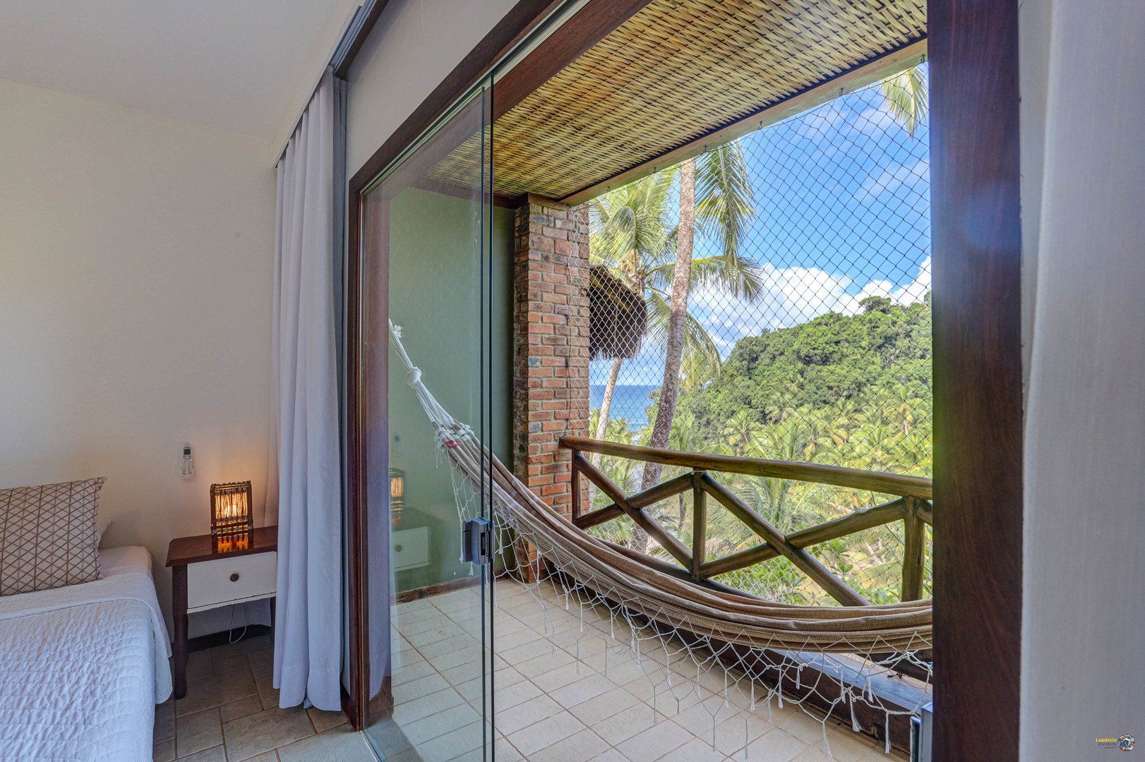 Quarto com uma grande janela, rede, cama e mesa de cabeceira.