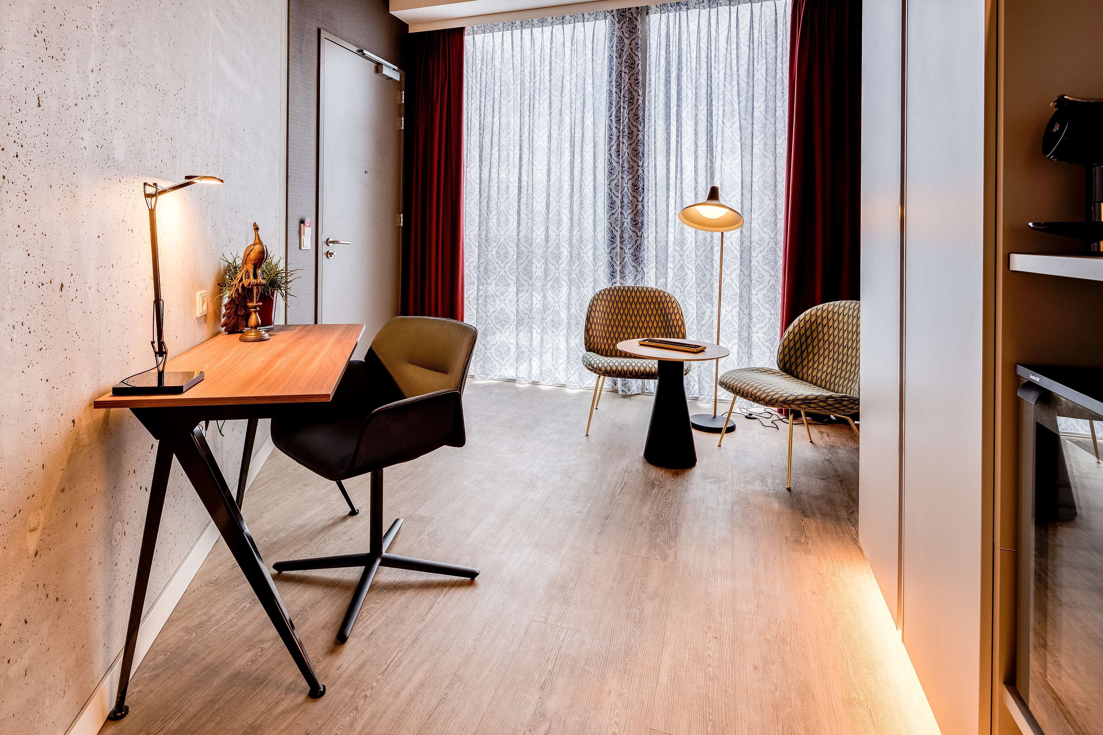 Quarto de hotel moderno com mesa de jantar de madeira, duas cadeiras, uma mesinha de apoio, abajur e uma janela grande com cortinas.