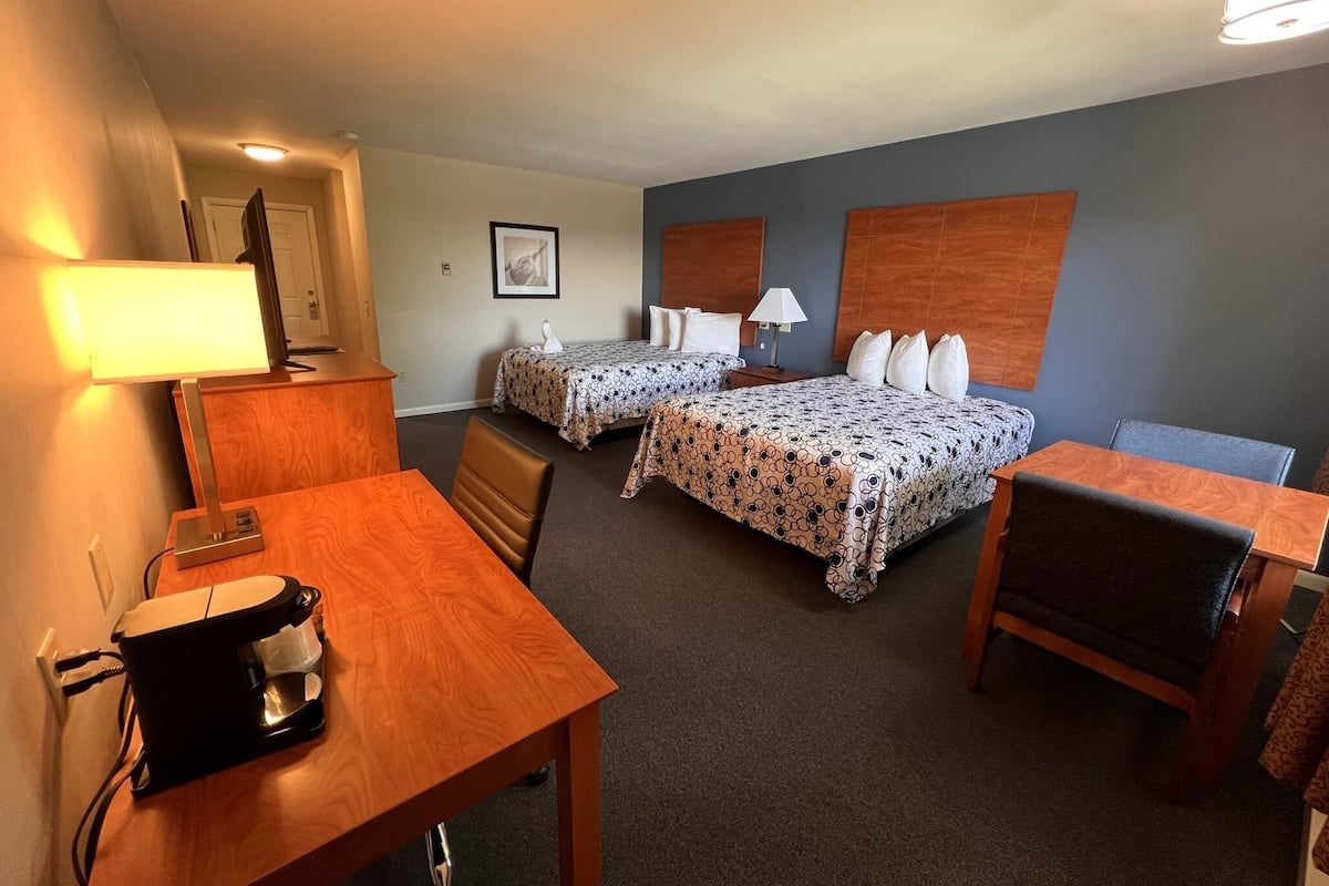 Quarto de hotel com duas camas, uma escrivaninha, uma cadeira, um abajur e uma cafeteira.