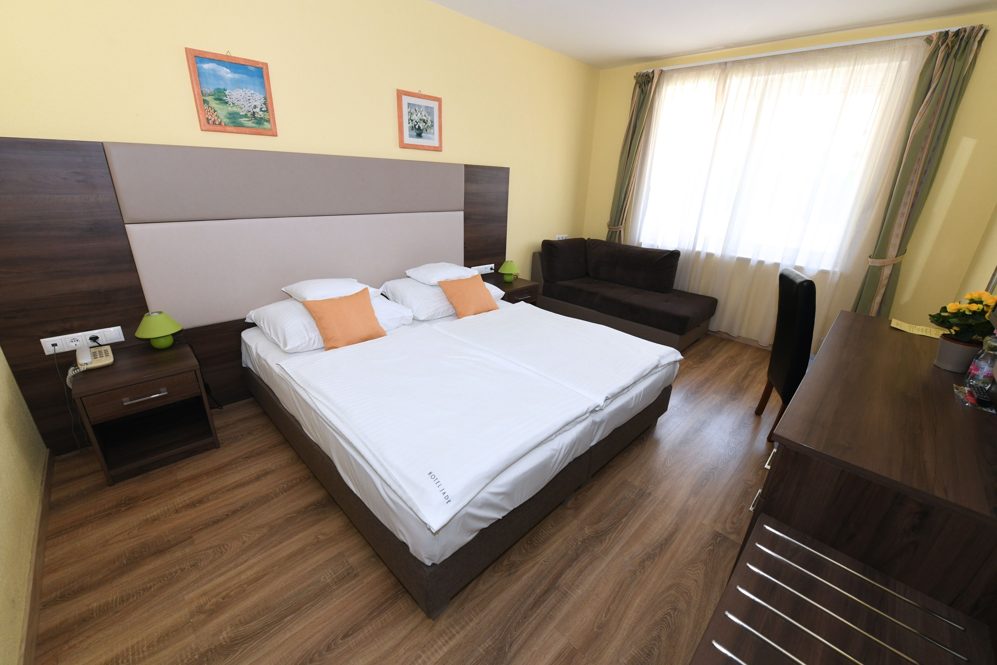 Quarto de hotel com uma cama grande, mesa de cabeceira com abajur, pequena escrivaninha, sofá e janela com cortinas.
